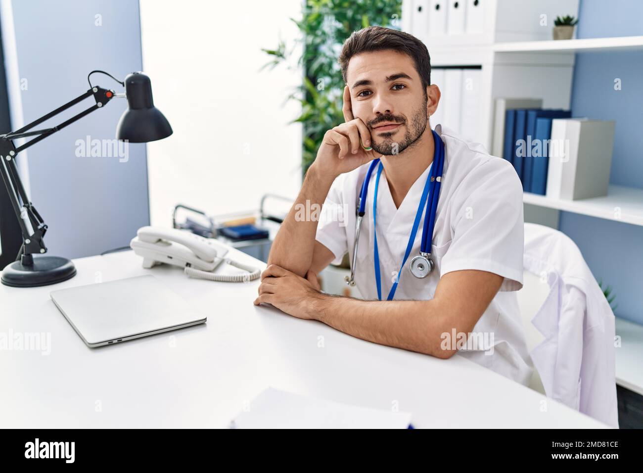 Young hispanic man wearing doctor uniform sitting on table at clinic ...