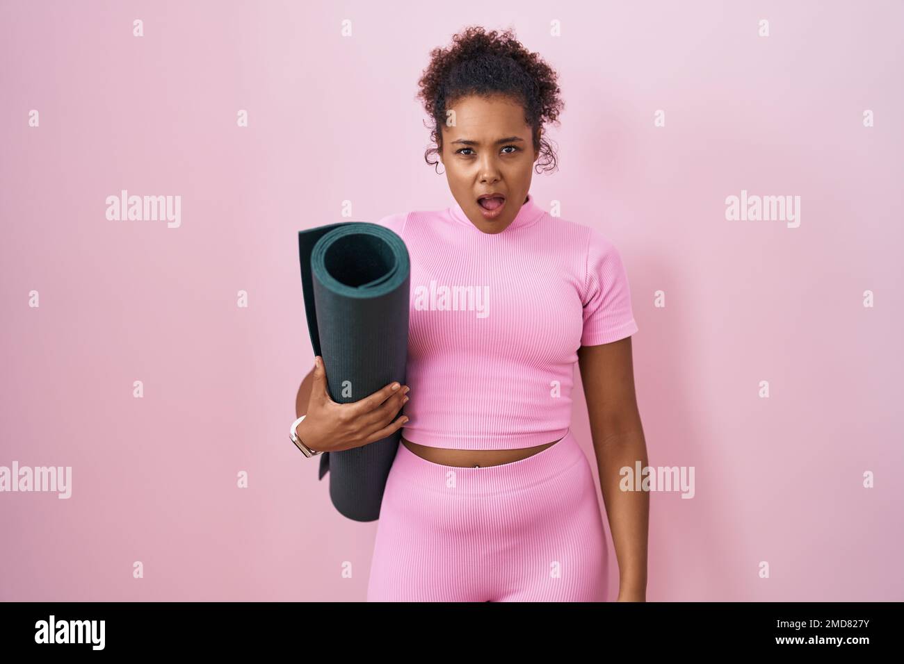 Young hispanic woman with curly hair holding yoga mat over pink ...