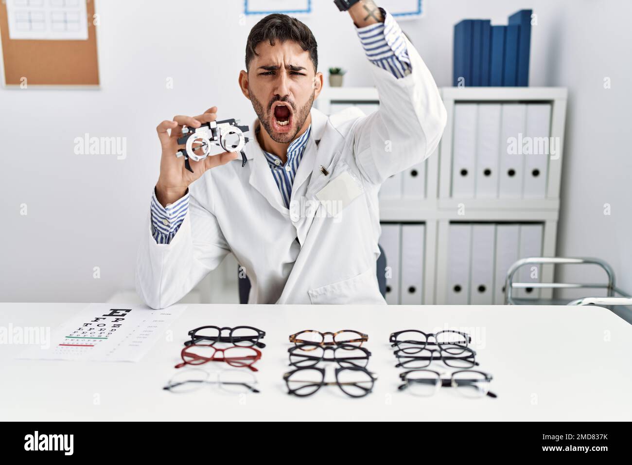 Young optician man holding optometry glasses angry and mad raising fist ...
