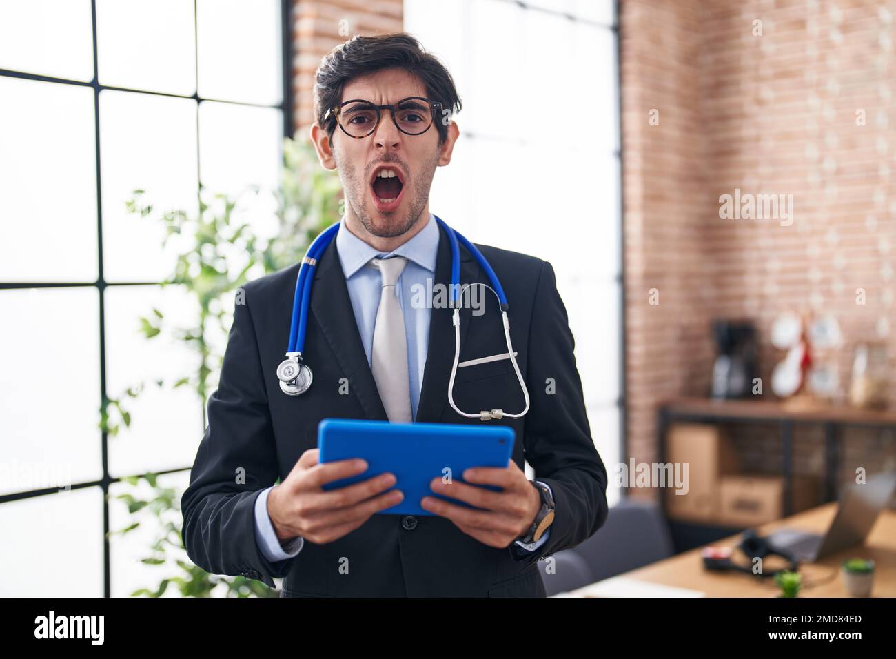 Young hispanic man wearing doctor stethoscope at the office angry and ...