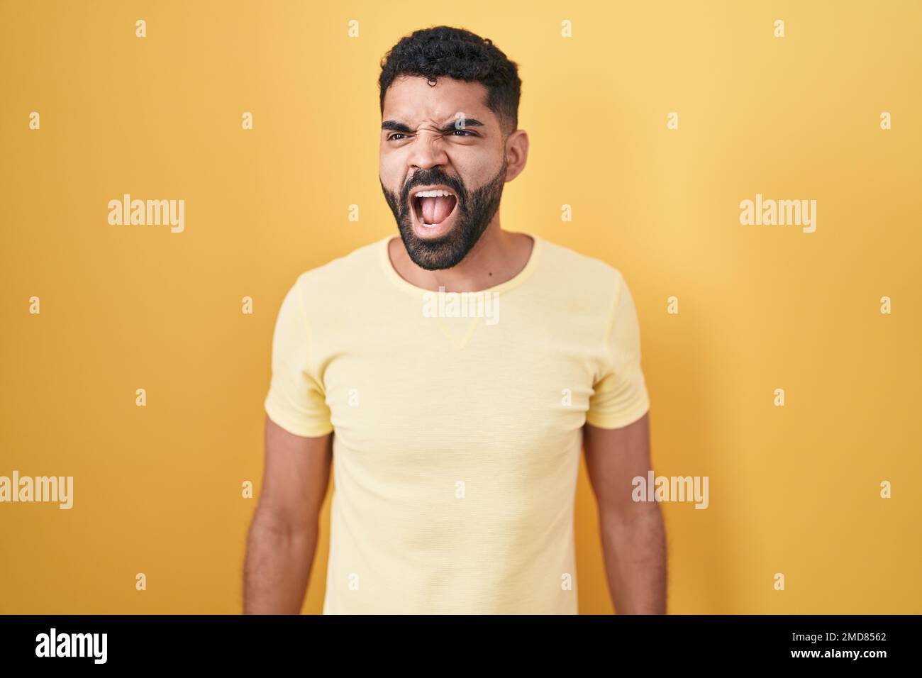 Hispanic man with beard standing over yellow background angry and mad ...