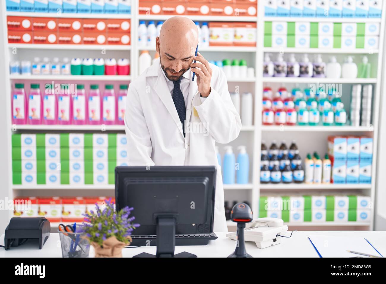 Young bald man pharmacist talking on smartphone using computer at ...