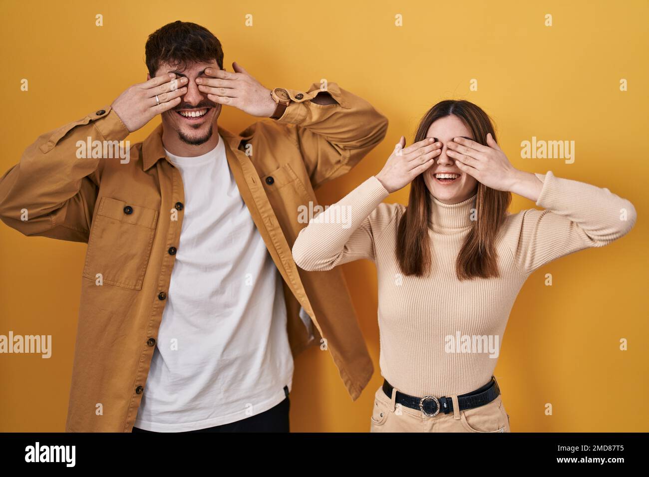 Young hispanic couple standing over yellow background covering eyes ...