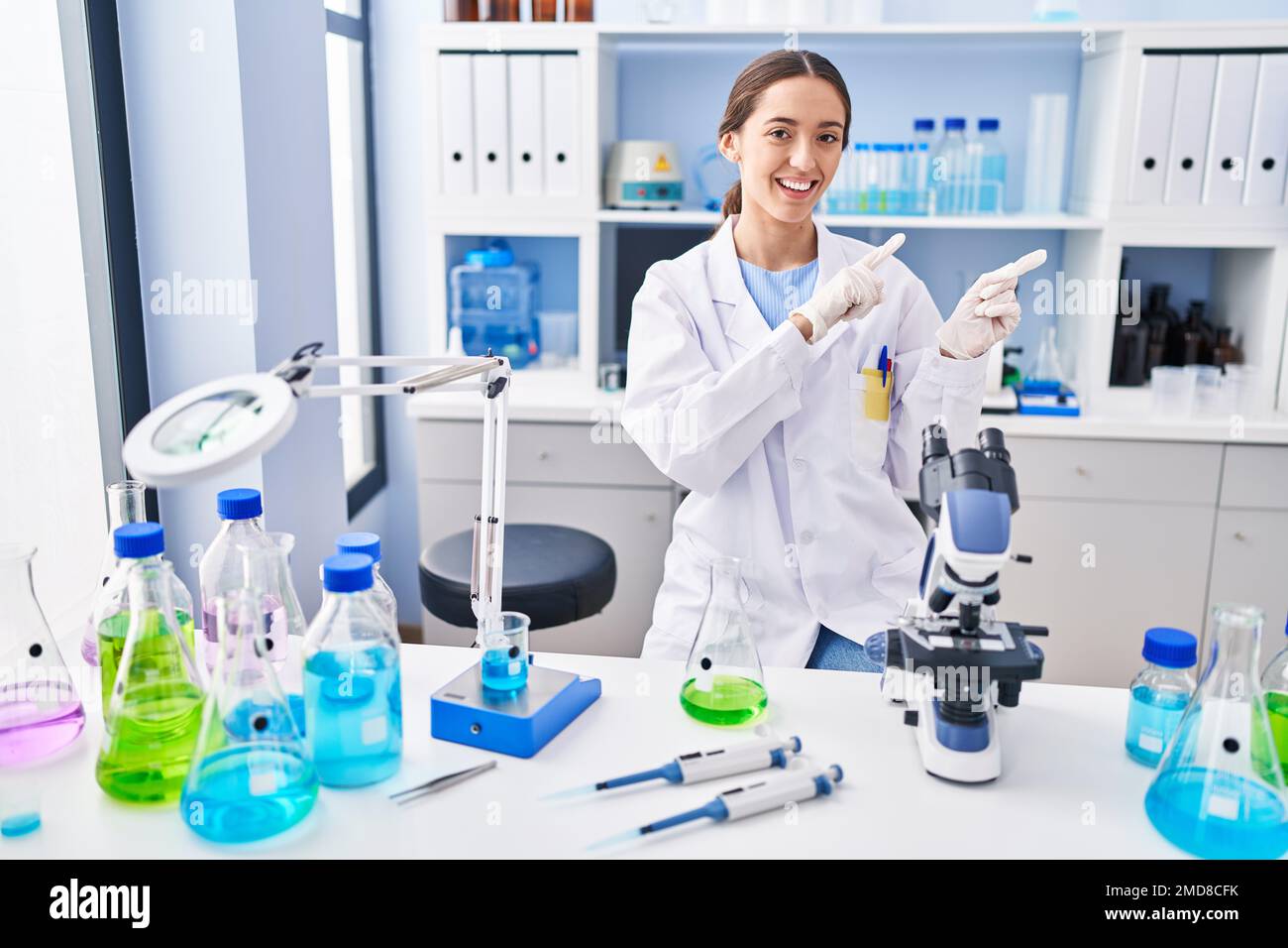Young brunette woman working at scientist laboratory smiling and looking at the camera pointing ...