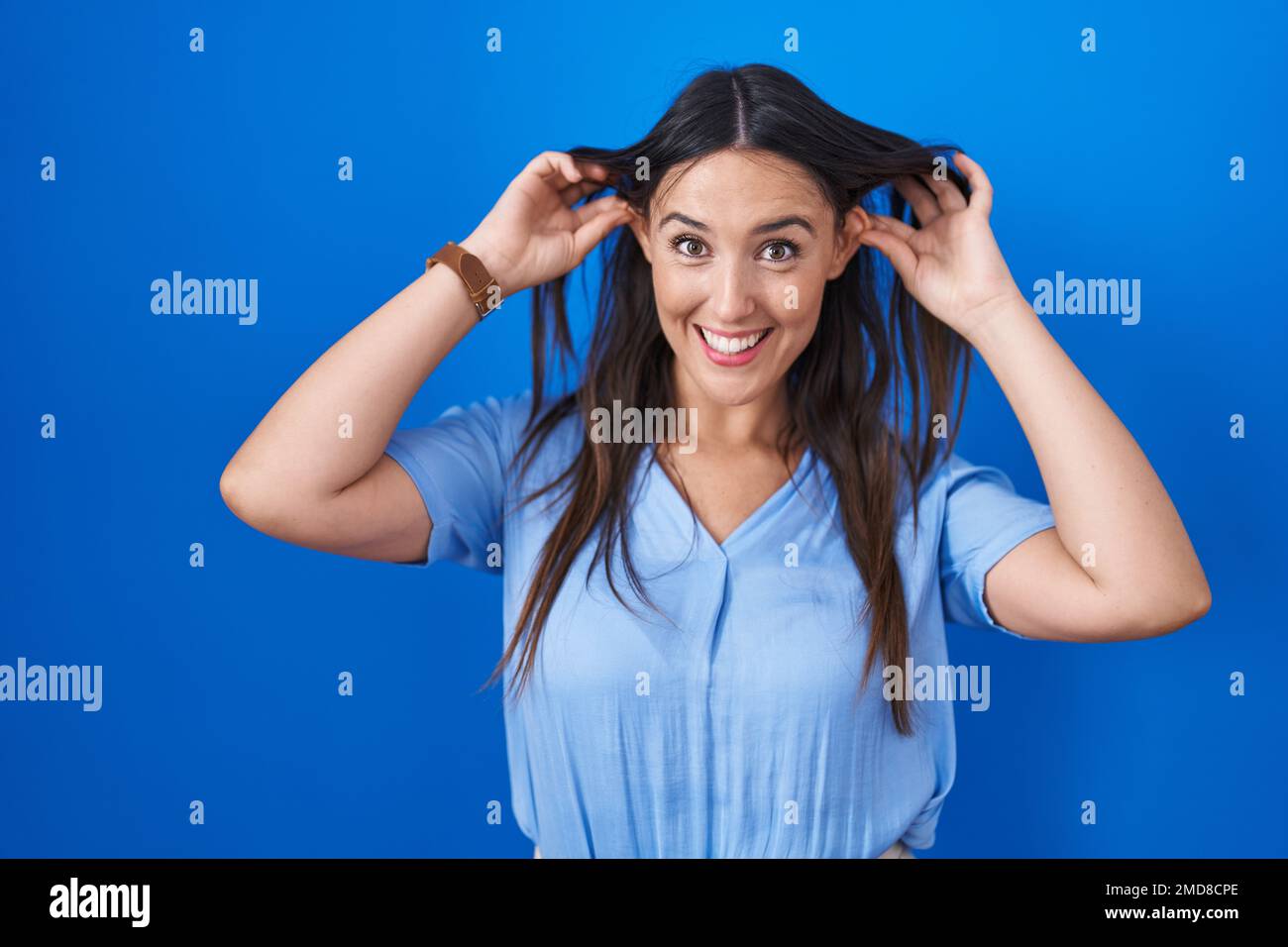 Young brunette woman standing over blue background smiling pulling ears ...
