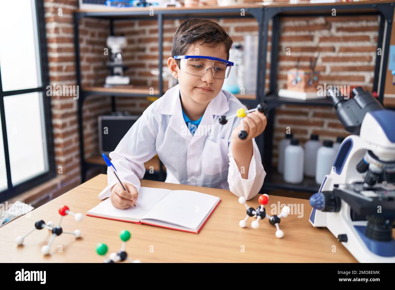 Adorable hispanic boy student writing on notebook holding molecules at ...