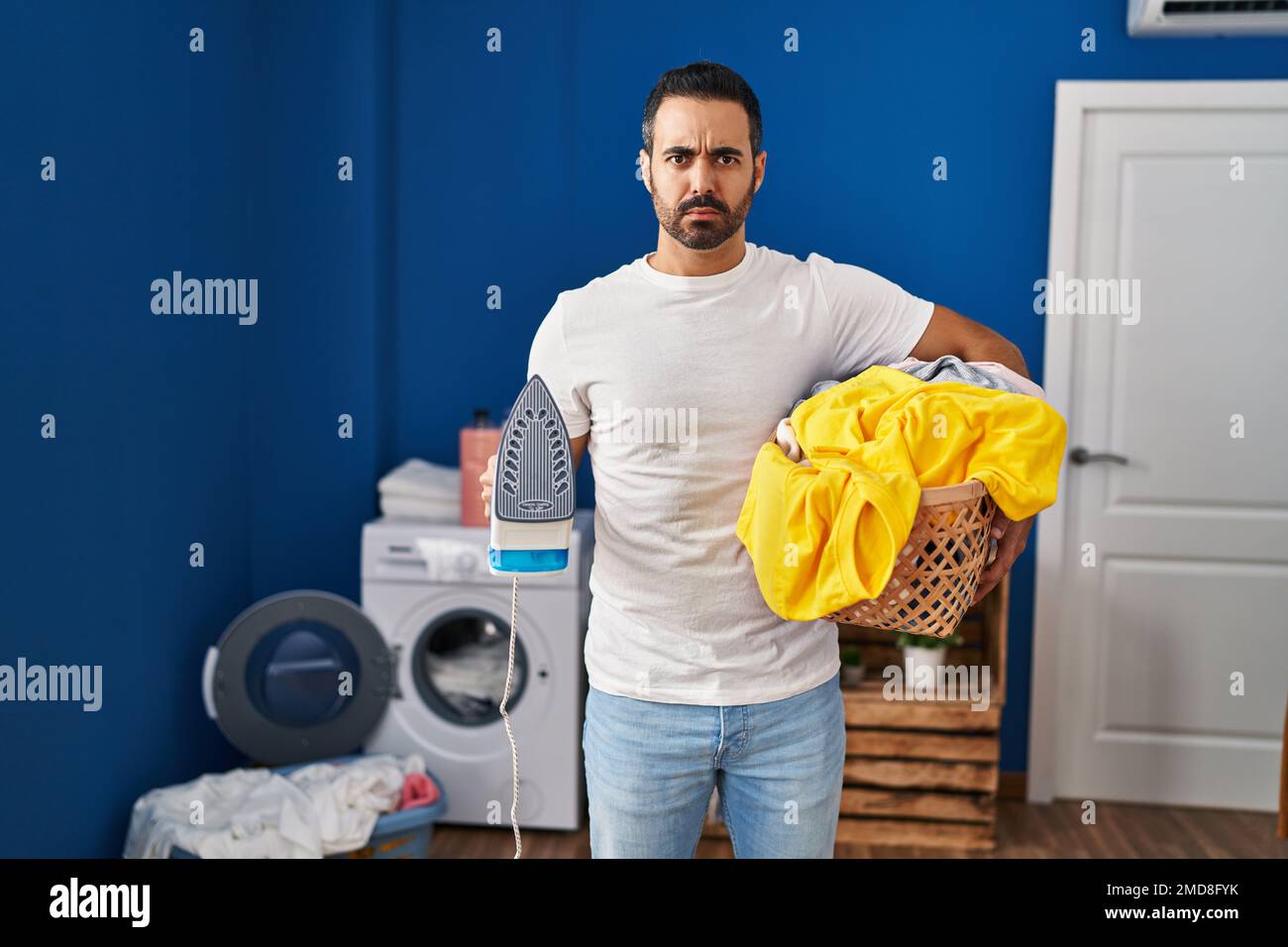Young hispanic man with beard holding iron and clothes at home skeptic ...