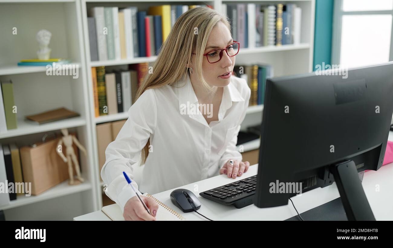 Young blonde woman student using computer writing on notebook at ...
