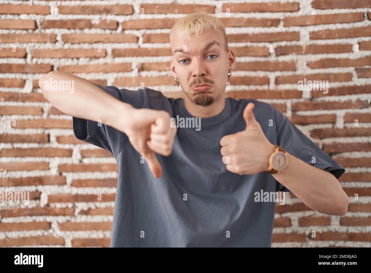 Young caucasian man standing over bricks wall doing thumbs up and down ...