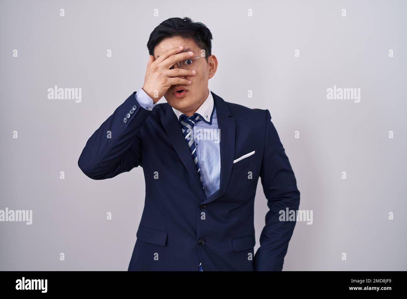 Young asian man wearing business suit and tie peeking in shock covering ...