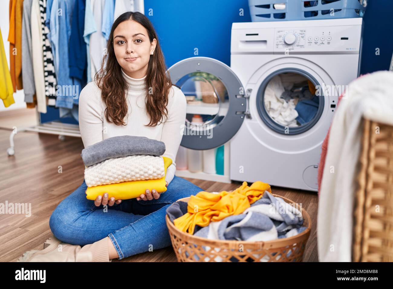 Young hispanic woman holding clean laundry puffing cheeks with funny face. mouth inflated with ...