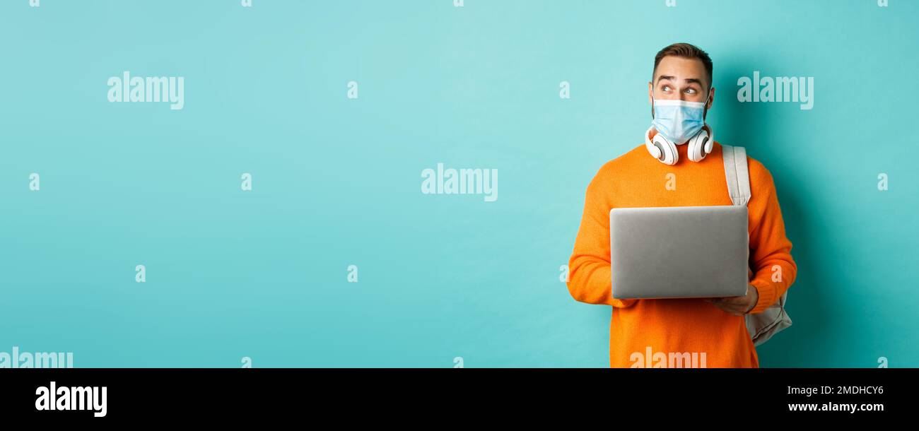 Happy young freelancer in medical mask, working on laptop, looking ...