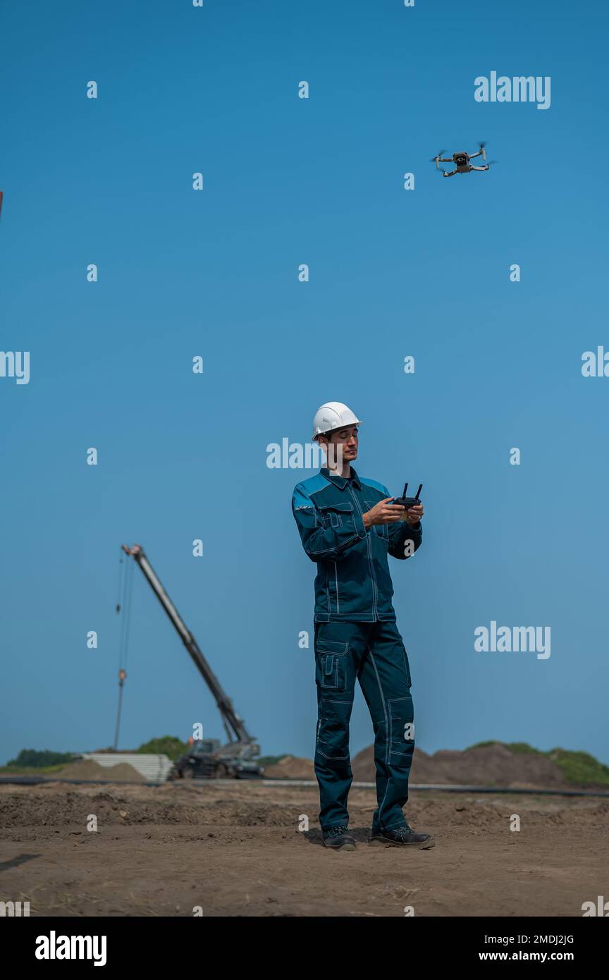 A man in a helmet and overalls controls a drone at a construction site ...