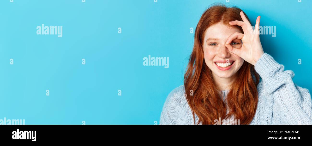 Headshot of cheerful redhead female model showing okay sign over eye ...