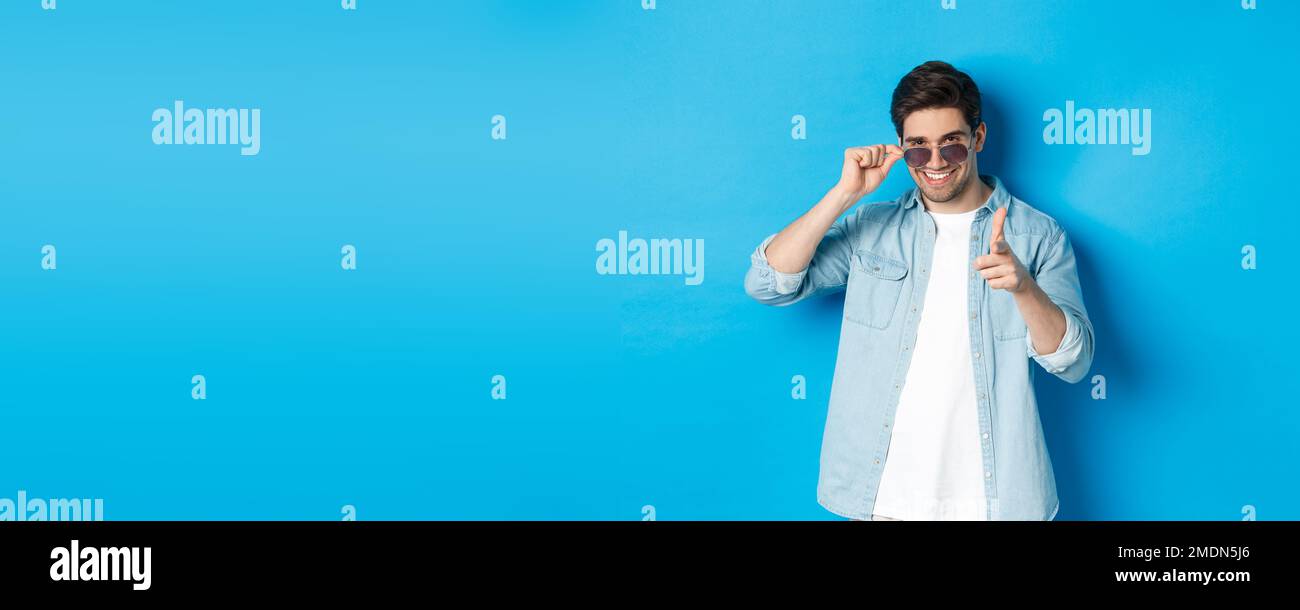 Handsome cheeky guy in sunglasses, smiling and pointing finger gun at ...