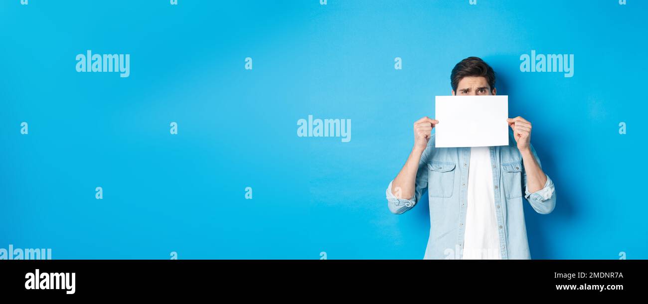 Shy and nervous guy hiding face behind blank piece of paper, peeking ...