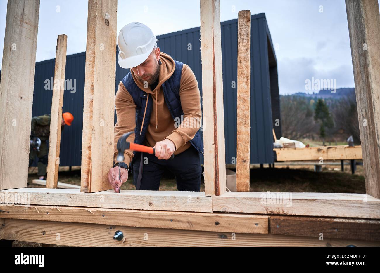 Man worker building wooden frame house on pile foundation. Carpenter hammering nail into wooden ...