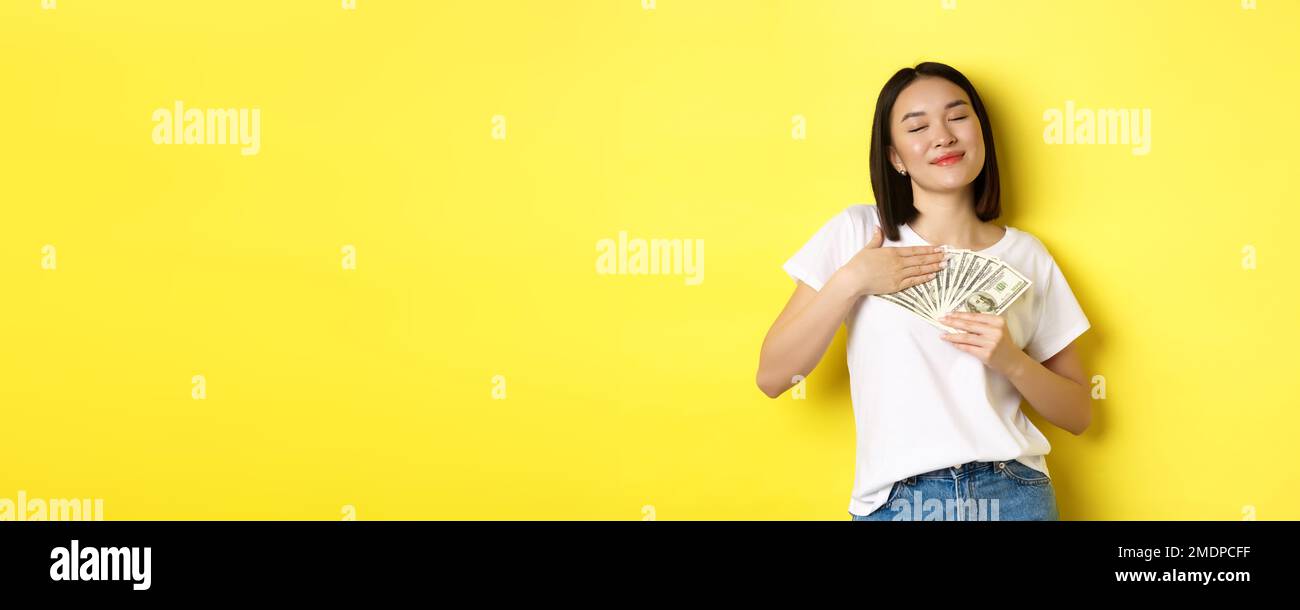 Beautiful asian woman love money, hugging dollars and smiling pleased ...