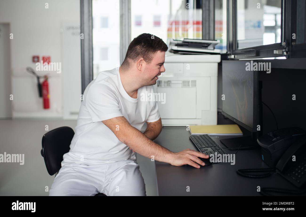 Young man with down syndrome working in hospital office, writing something on computer. Concept ...