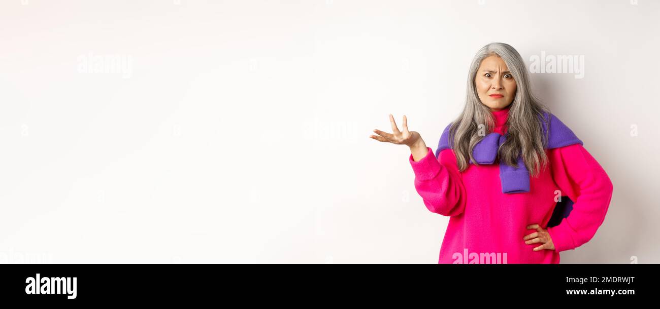 Angry and confused asian senior woman spread hand sideways and staring ...
