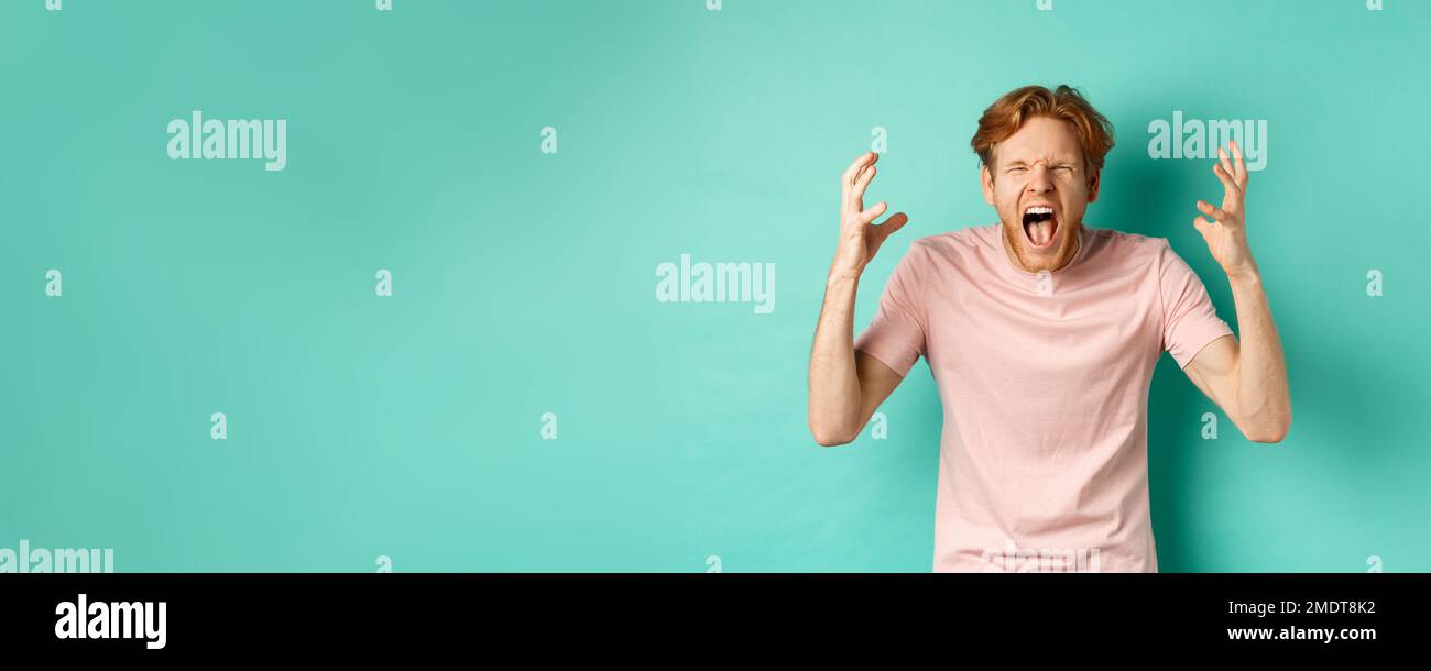 Angry young redhead man screaming distressed, shaking hands and looking ...