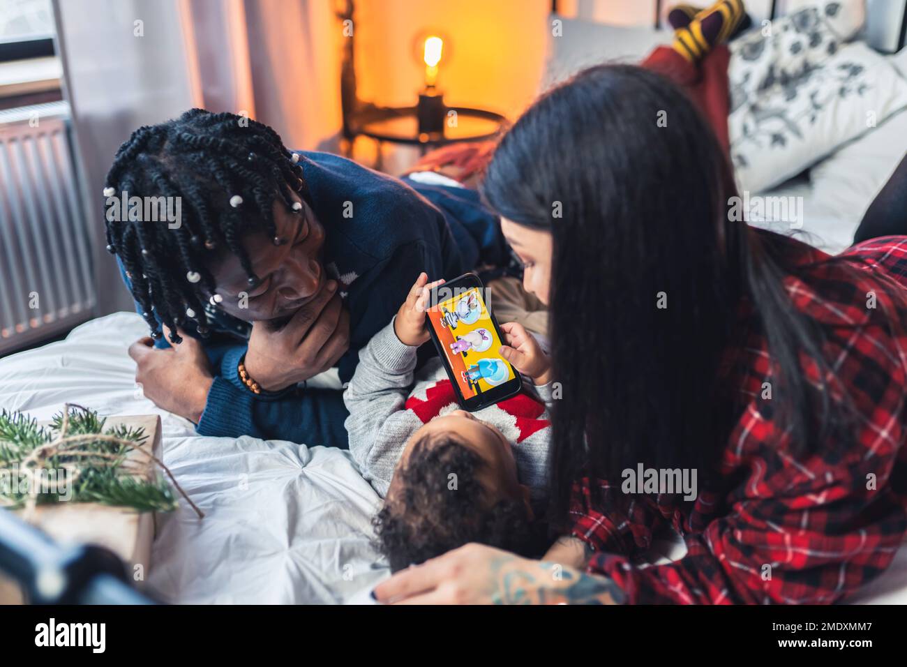 Multiethnic parents with their toddler son playing on the phone in bed ...