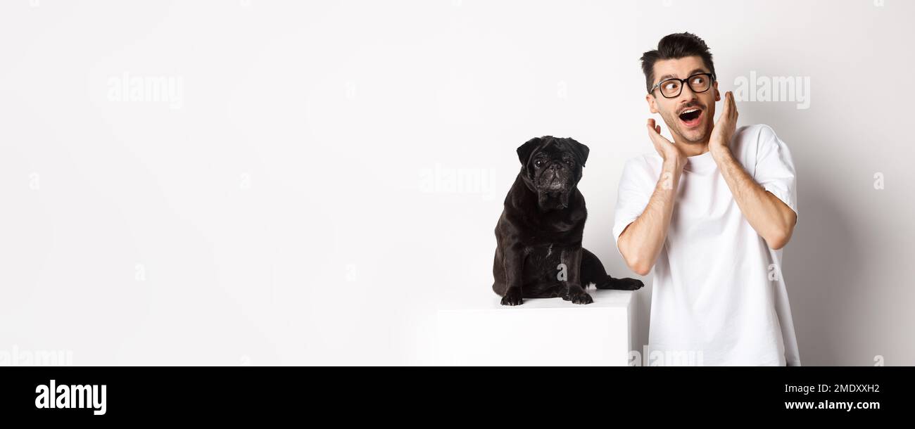 Amazed young man standing with cute black puppy, staring at upper right ...