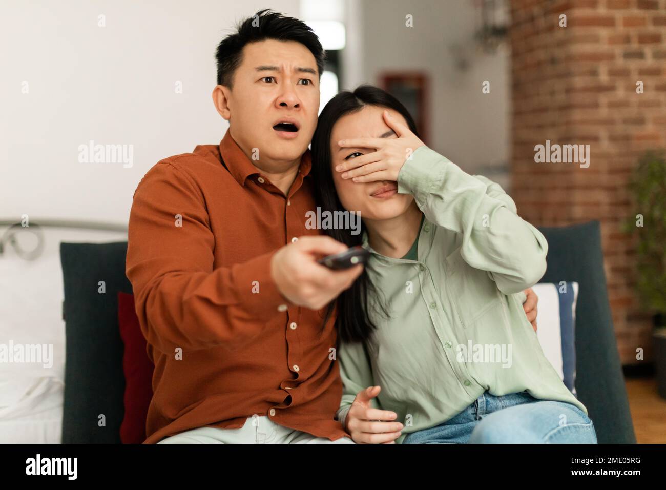 Scared asian woman and man with remote control switching channel on tv ...