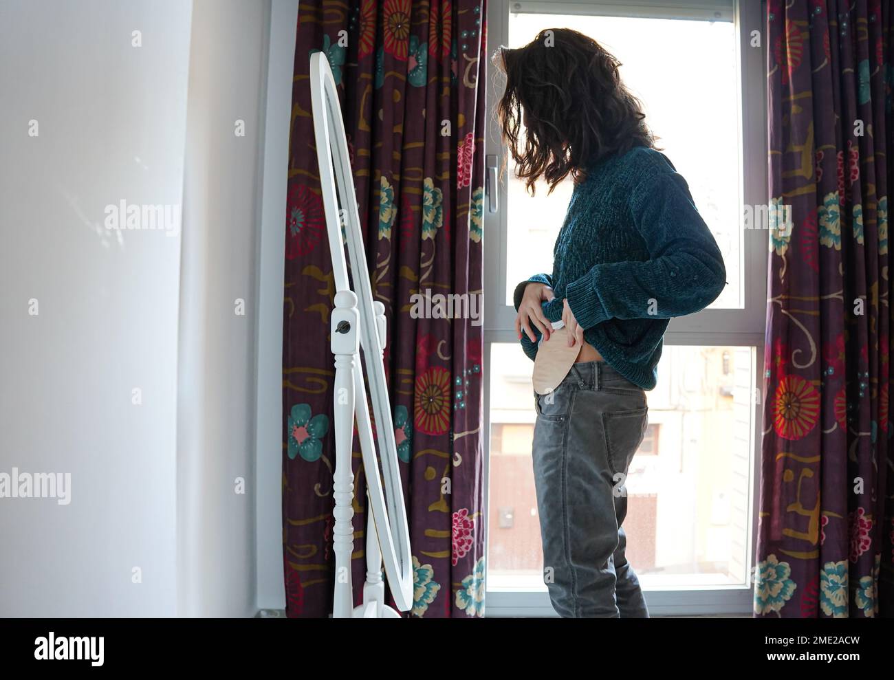 woman looks at her ostomy bag in the mirror at home. colon cancer ...
