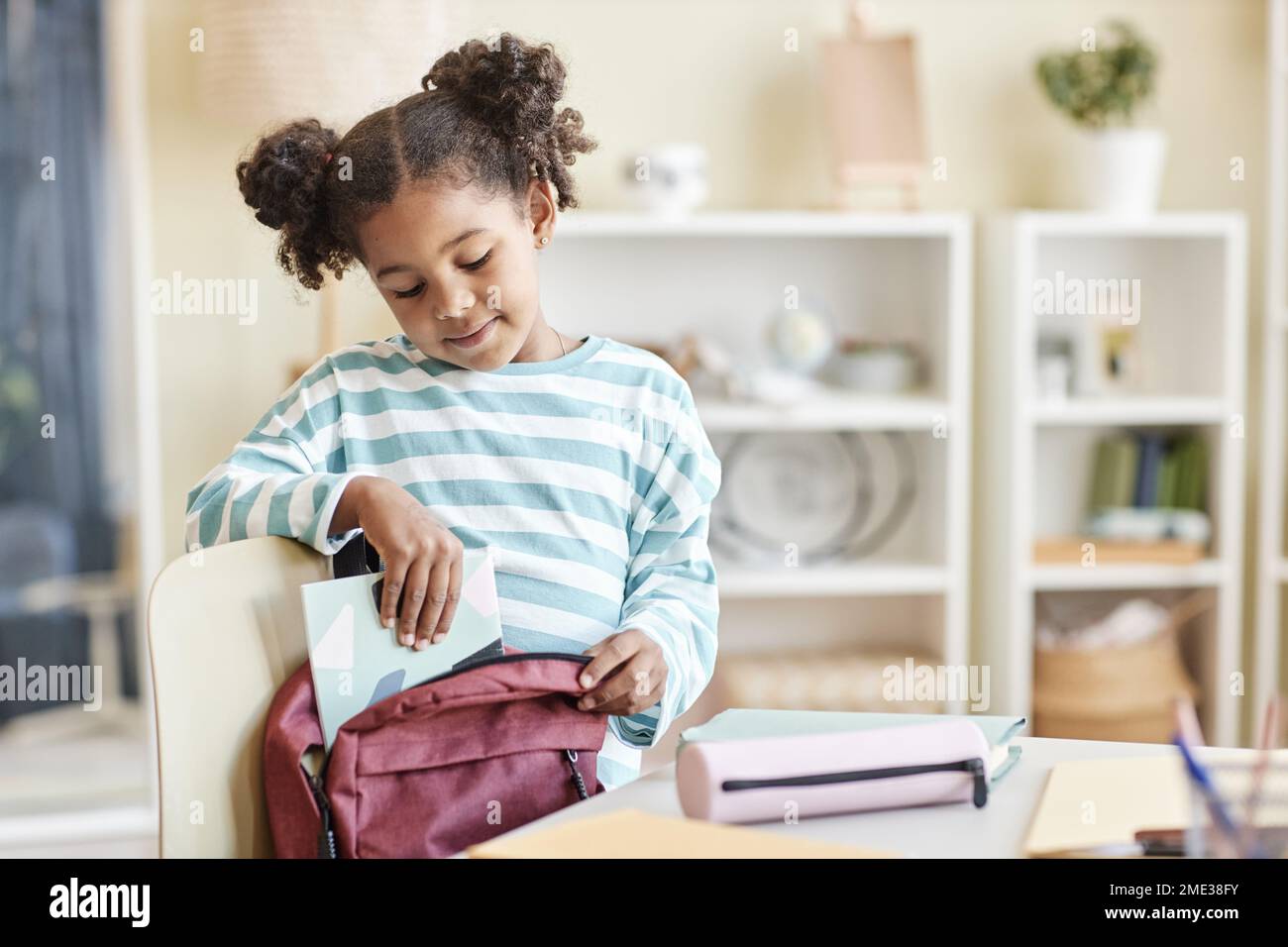 Minimal portrait of cute black girl packing backpack for school in ...