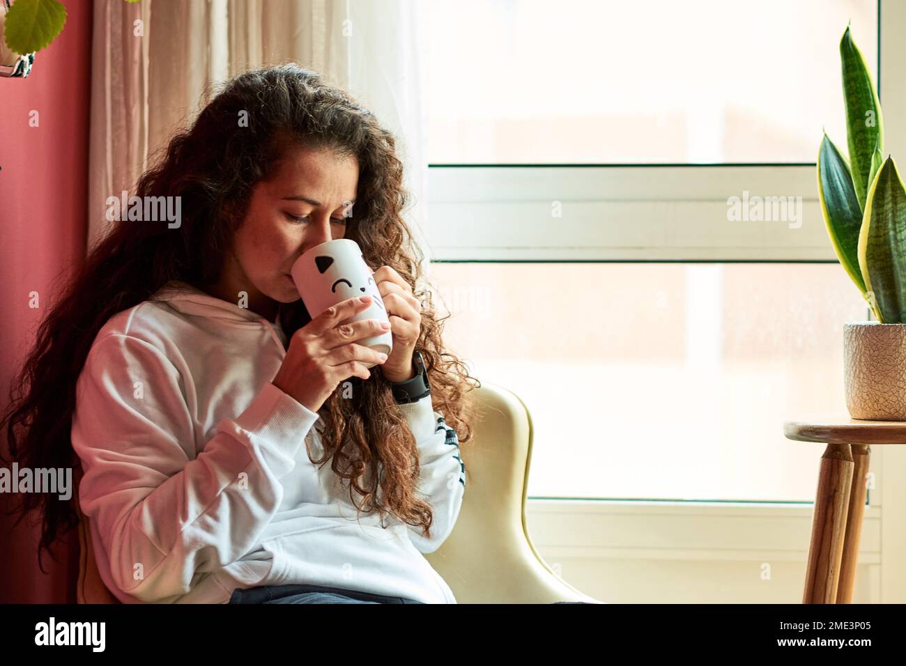 Woman relaxing in a armchair drinking a cup of tea indoors surrounded ...