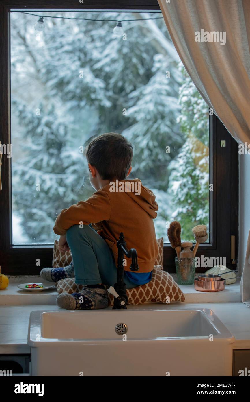 Boy sitting on window sill looking out in kitchen at home Stock Photo ...