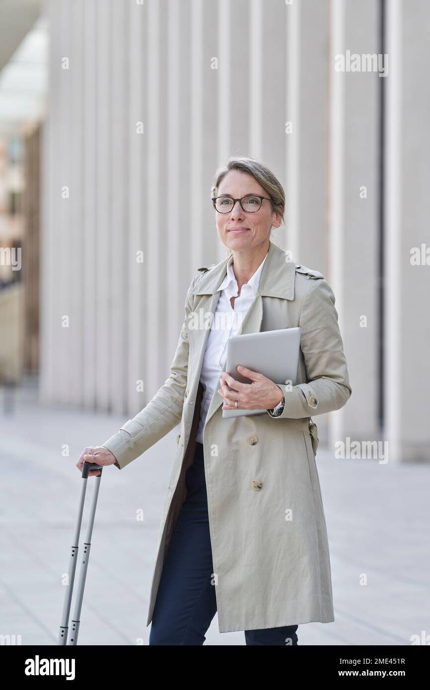Mature businesswoman with wheeled luggage standing in city holding ...