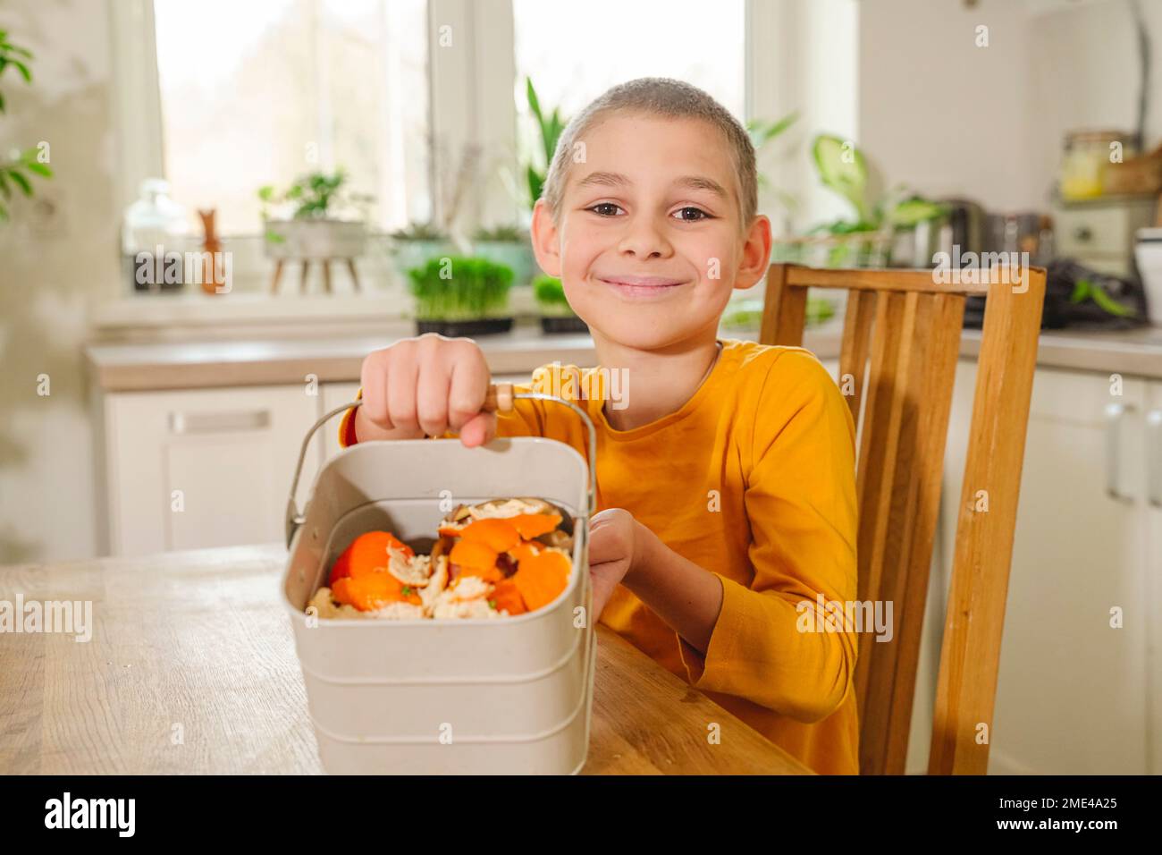 Smiling boy showing mandarin peels in compost bucket at dining table ...
