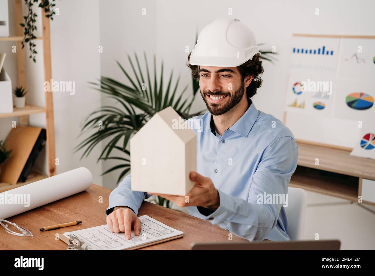 Smiling architect with blueprint holding house model at desk in office ...