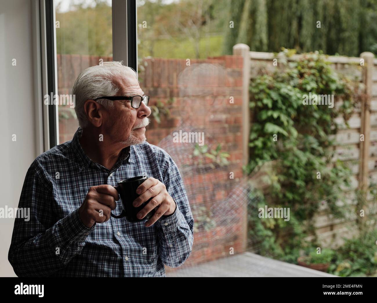 Pensive senior man with cup of coffee looking out of the window at home ...