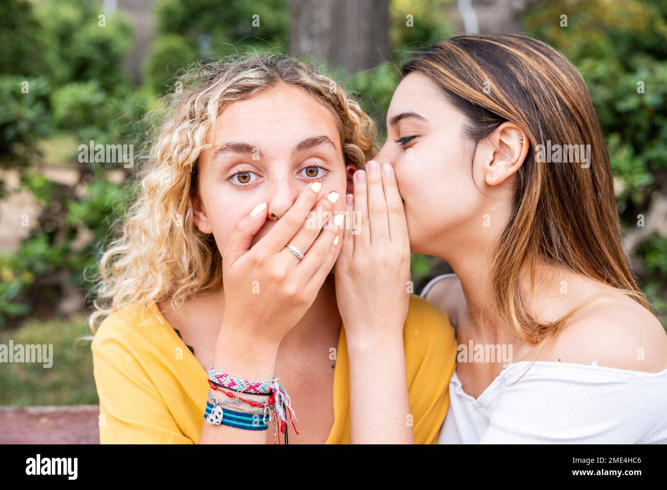 Woman whispering into shocked friend's ear in park Stock Photo - Alamy