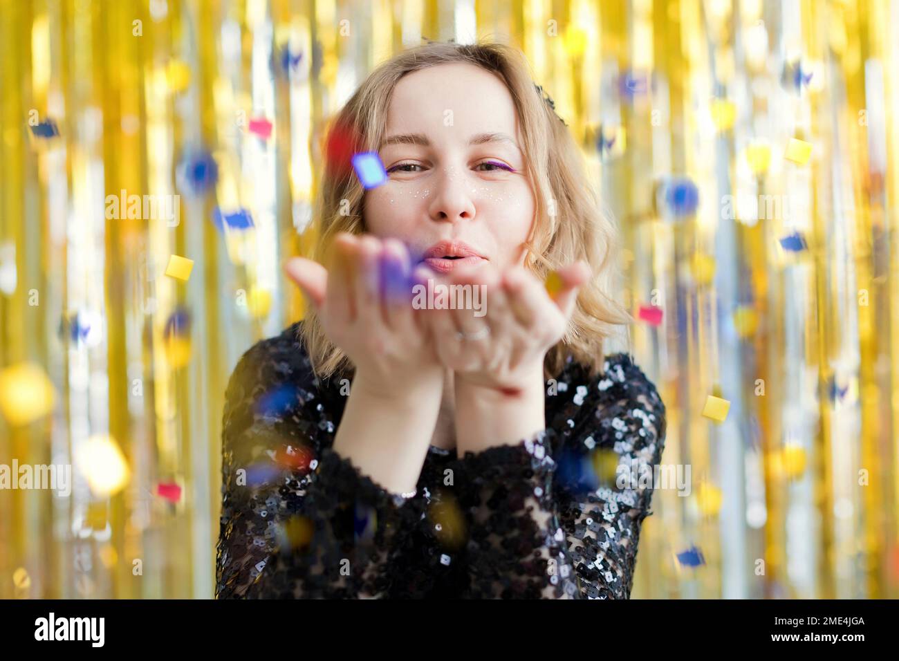 Woman wearing black dress blowing confetti in front of golden tinsel ...