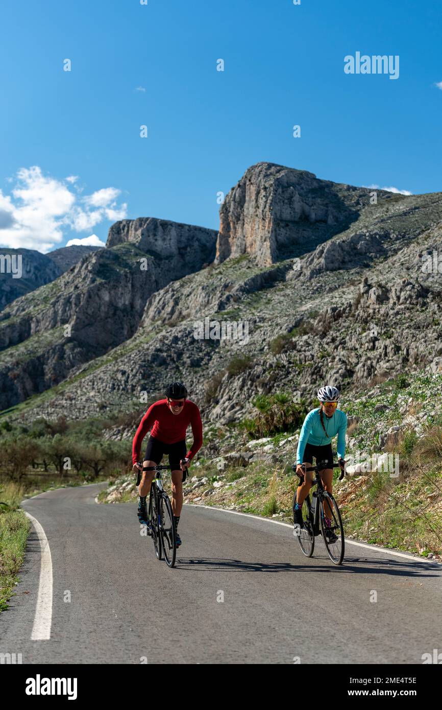 Mature cyclists together riding bicycles on road in front of rocks ...