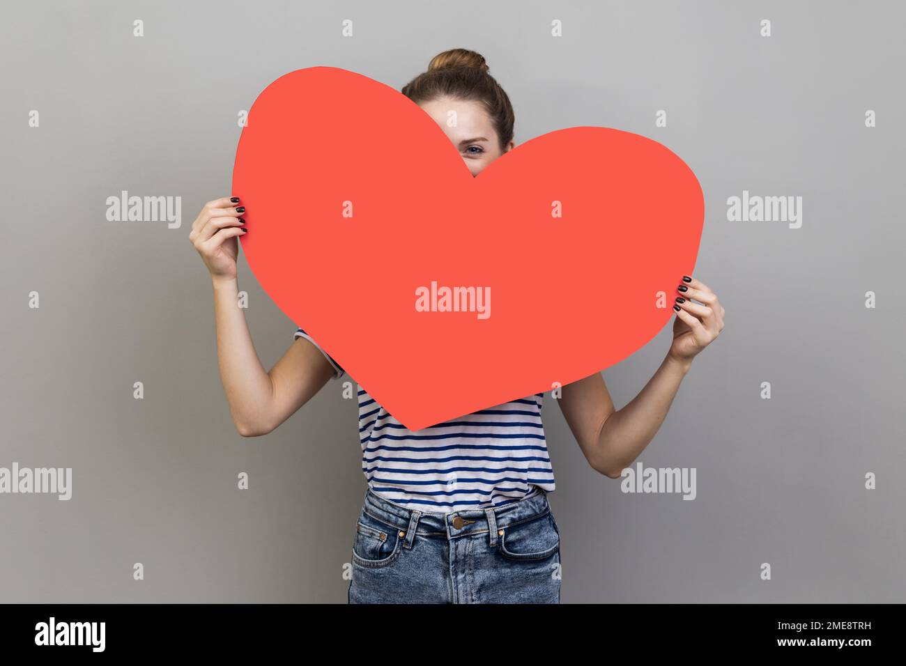Portrait of woman hiding face behind big red heart and looking at ...