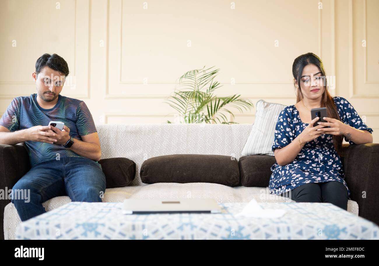 Young Indian couple busy using mobile phone while sitting at sofa in ...