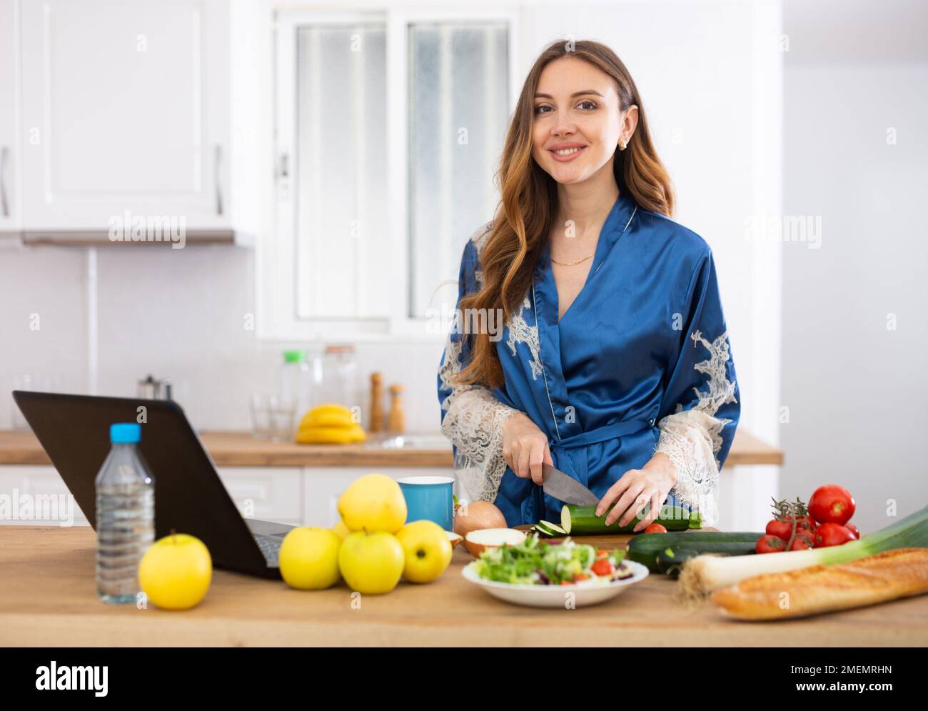 Young woman in blue robe cooking and watching TV series on laptop Stock ...