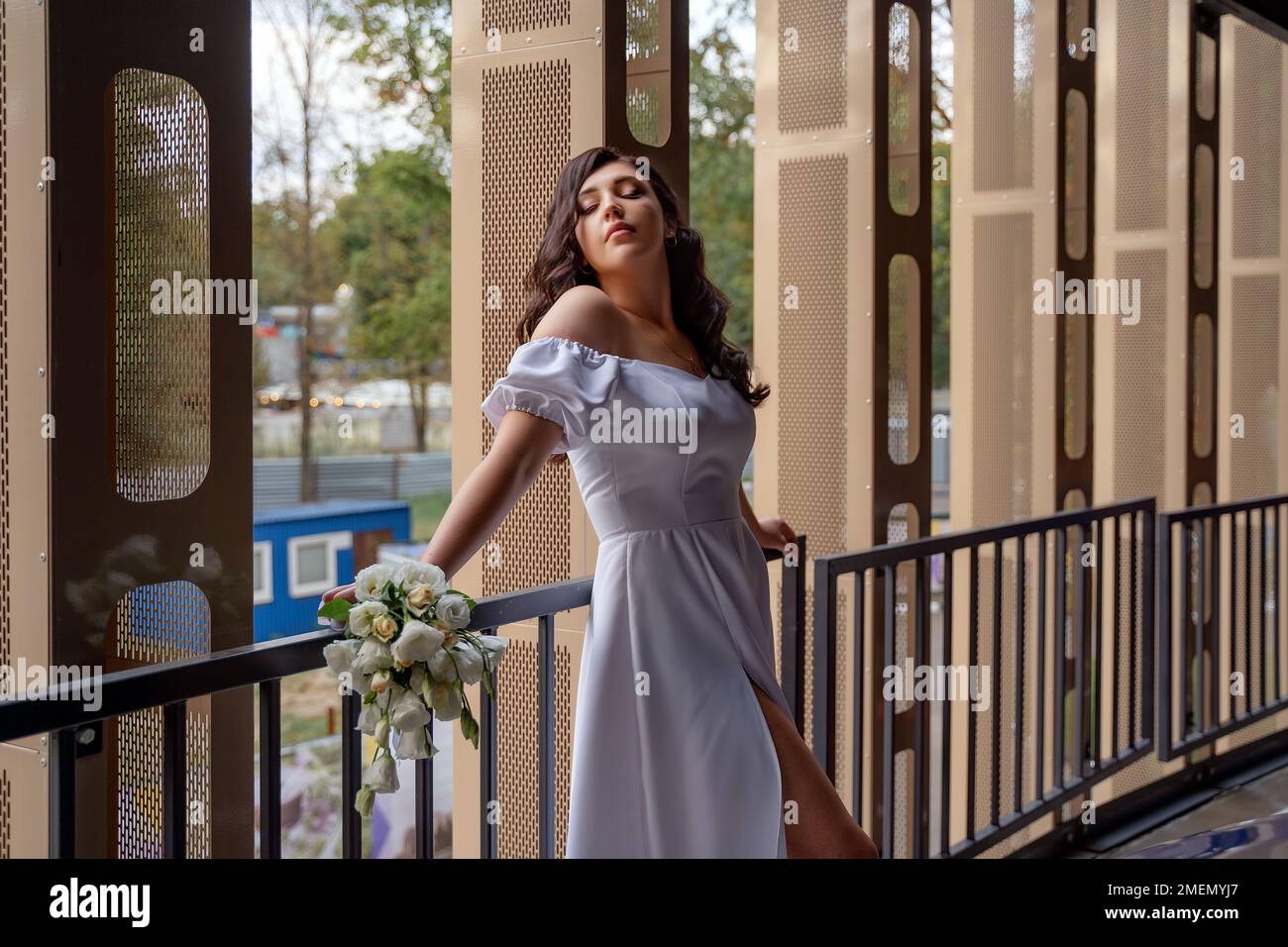 Bride in chic dress stands leaning on the railing in the parking lot ...