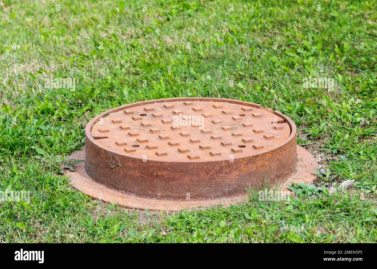 Rusty man hole in grass. Top is covered with square bumps. Focus on ...