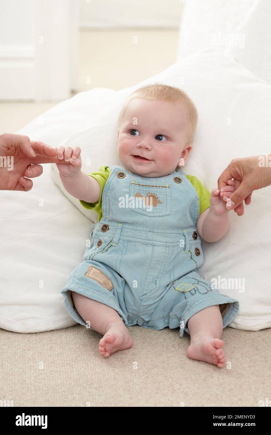 Two people holding hands of baby boy (17 weeks) as he sits supported by ...