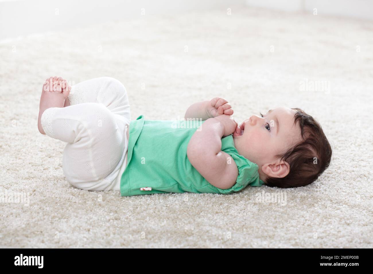Baby girl (6 months) lying on back on floor with her hand to her mouth ...