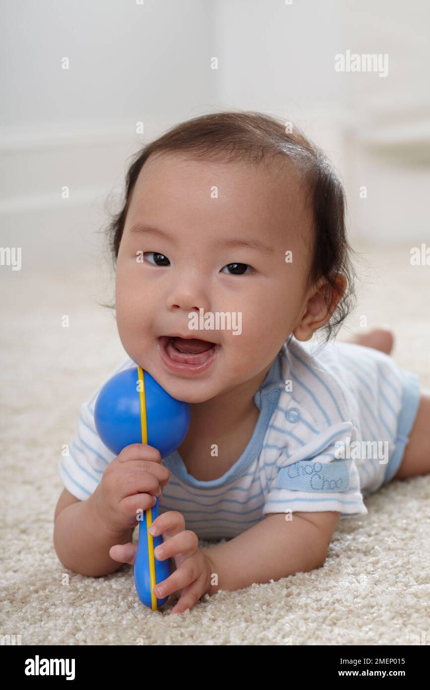 Baby boy (6 months) lying on front on carpet smiling and holding a ...
