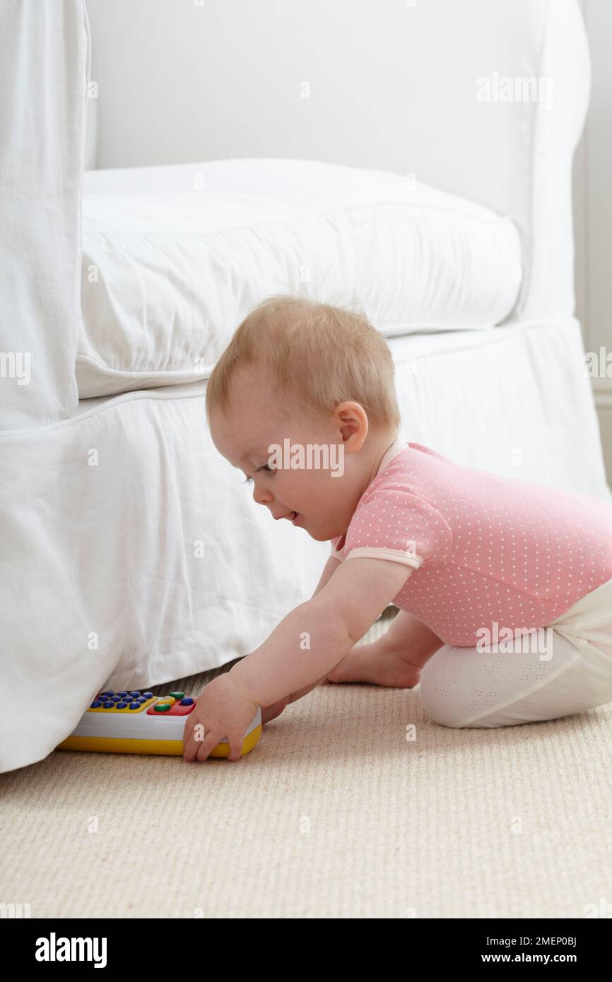 Baby girl (9 months) kneeling on carpet pushing toy phone under sofa ...