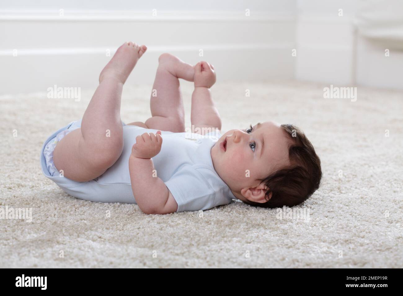 Baby girl (6 months) lying on back on floor with her legs up holding ...