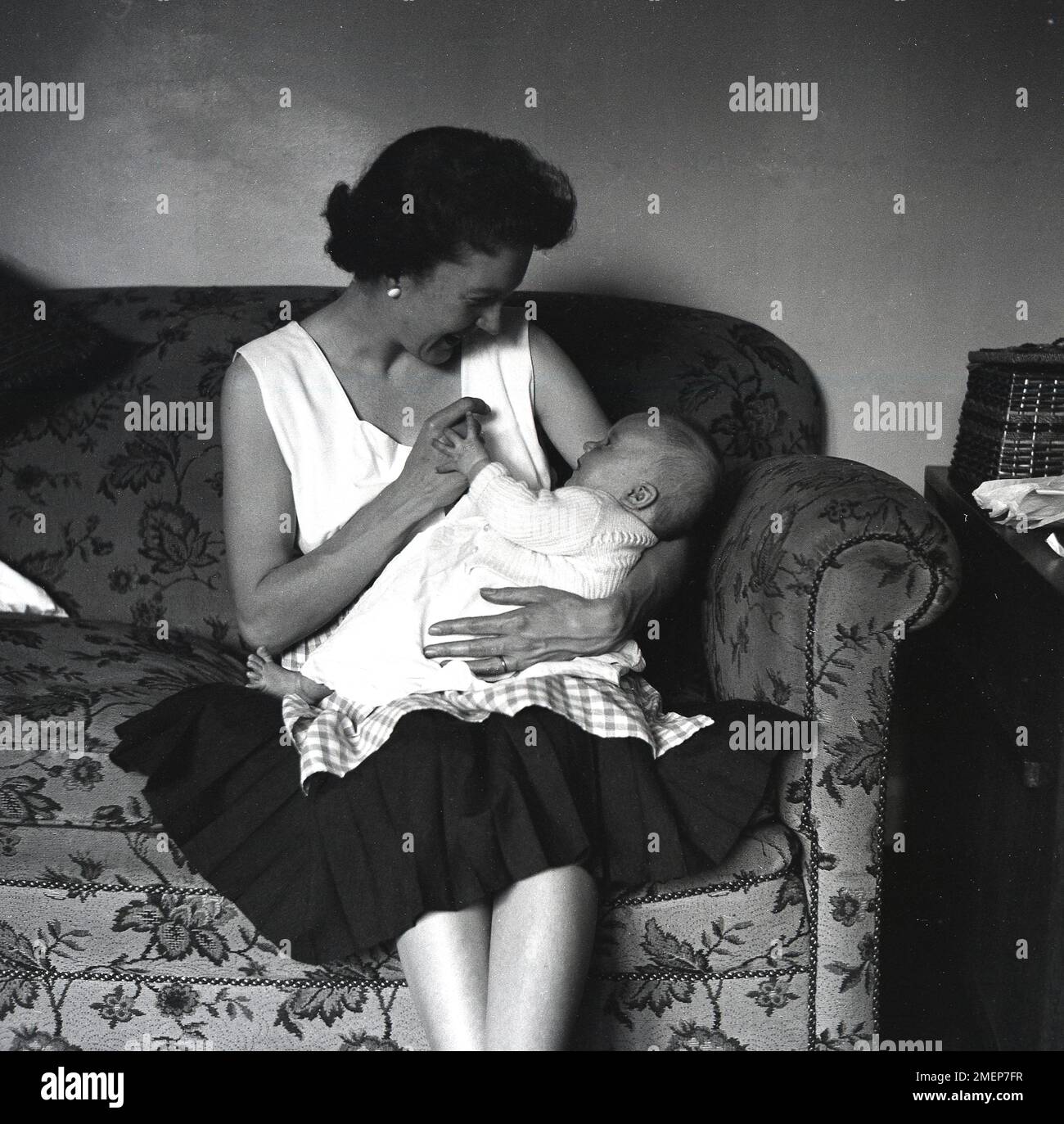 1950s, historical, inside a room, a mother sitting on a sofa cradling ...